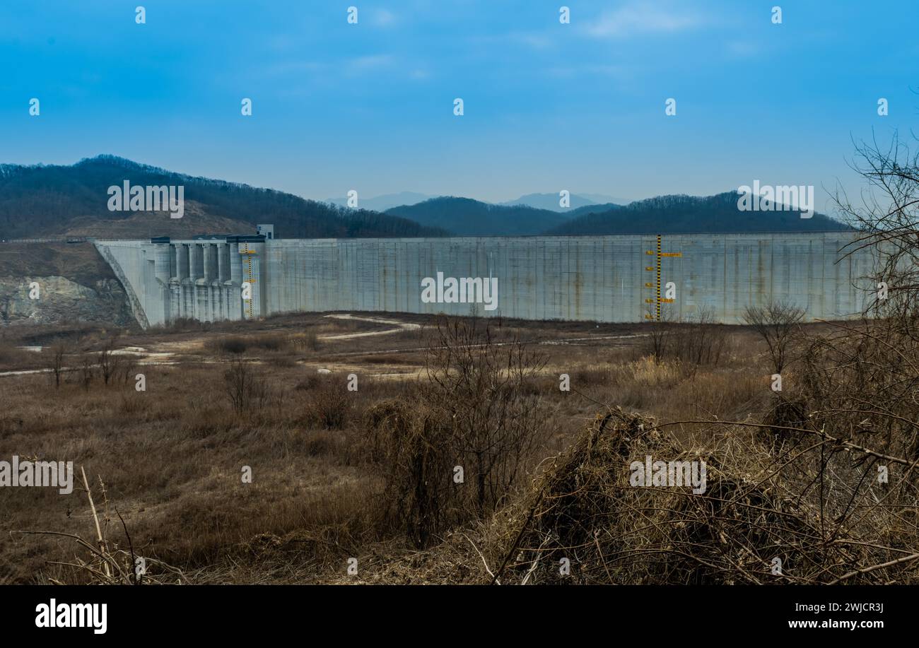 Field of dry grass and trees in front of concrete dam with yellow and ...