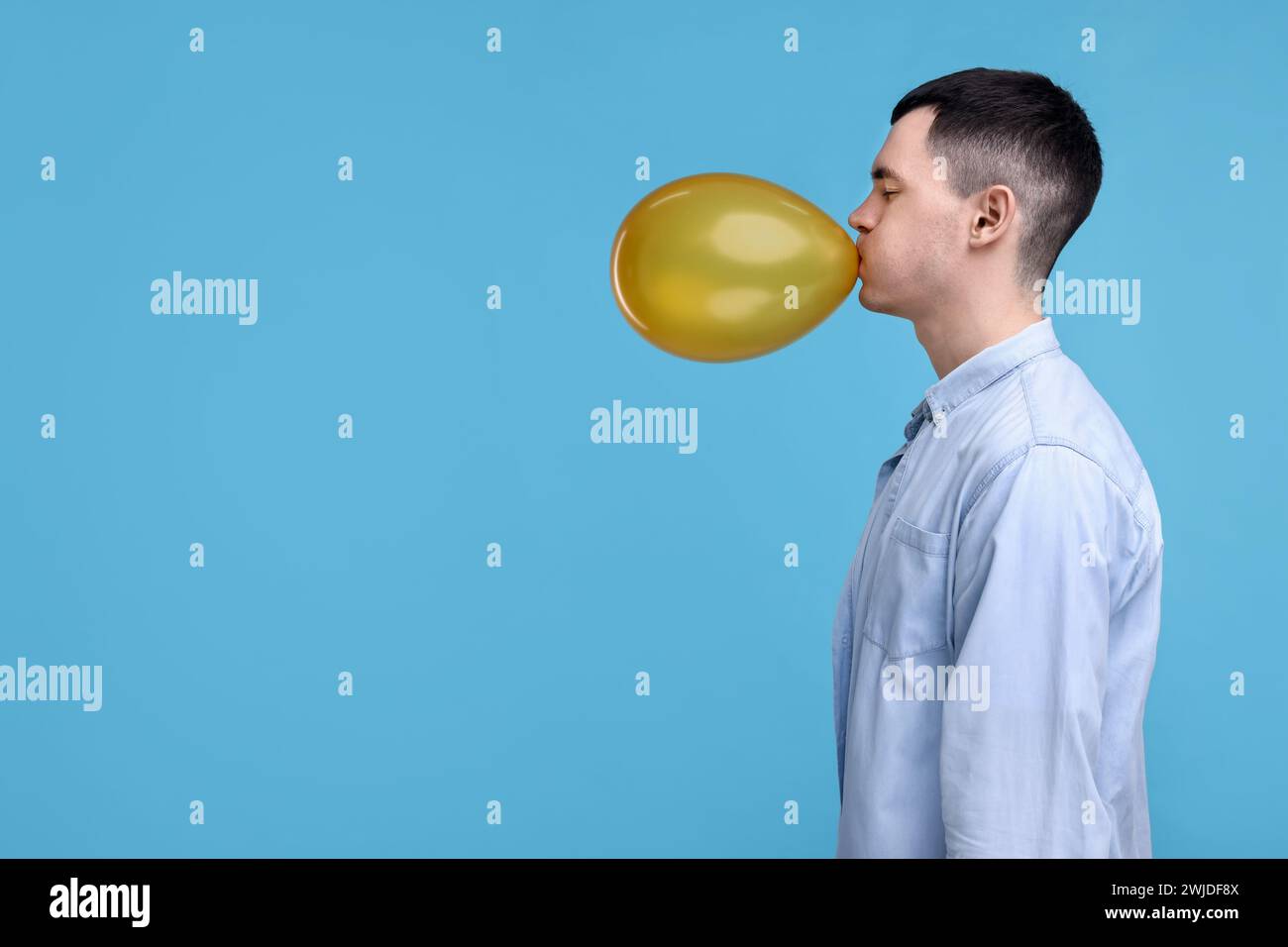Young man inflating golden balloon on light blue background. Space for ...