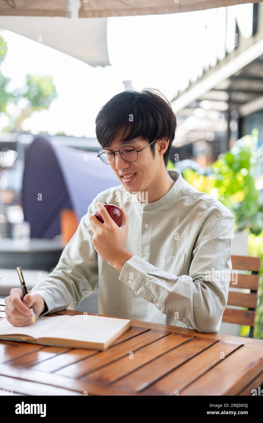 A handsome young Asian man is eating an apple while keeping his diary or writing down his ideas ...