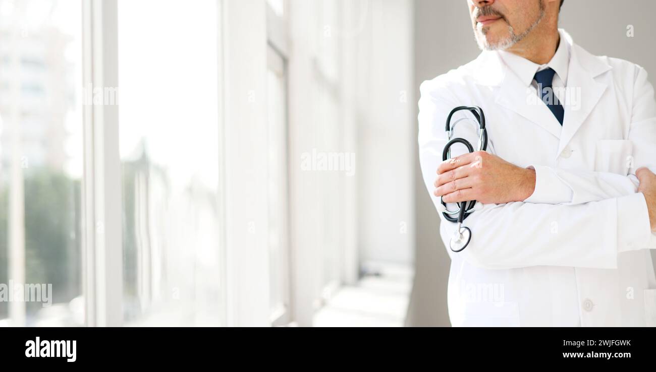 A thoughtful senior doctor with a stethoscope, standing by a window in ...
