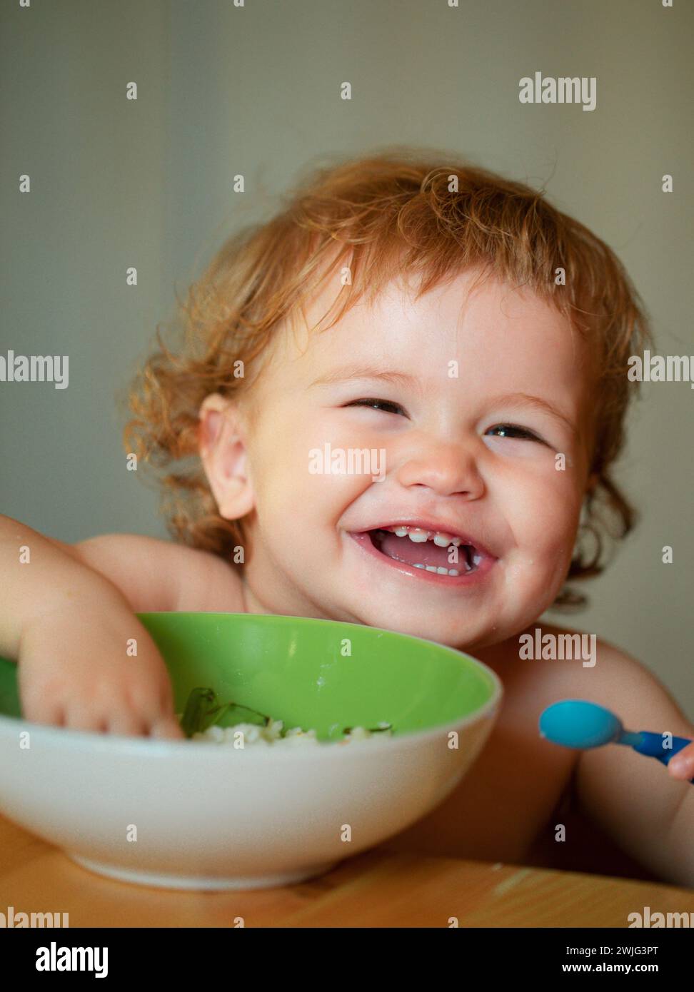 Funny baby eating food himself with a spoon on kitchen. Smiling baby ...