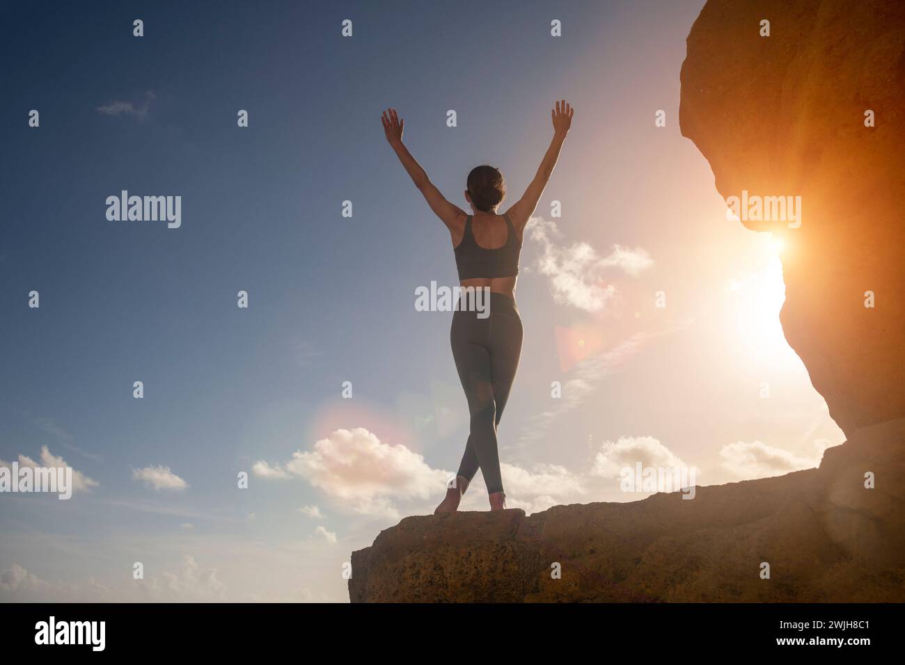 sporty woman standing on top of a rock at sunrise with her arms raised ...