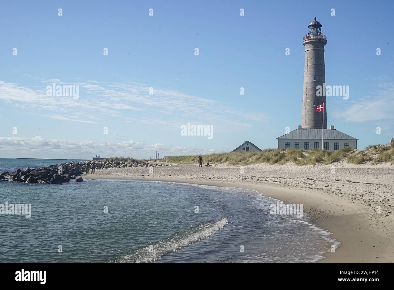 A lighthouse next to a beach under a blue sky with a Danish flag at the ...