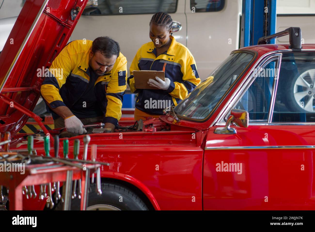 Two mechanics dressed in uniform fix a car together in a garage full of ...