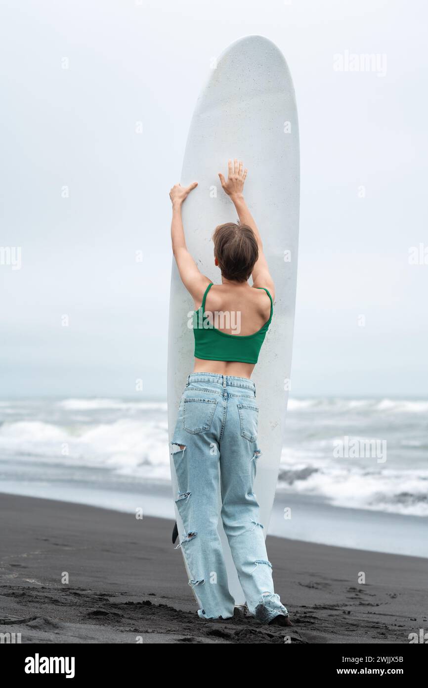 Rear view of woman surfer arms raised and holding surfboard vertically ...