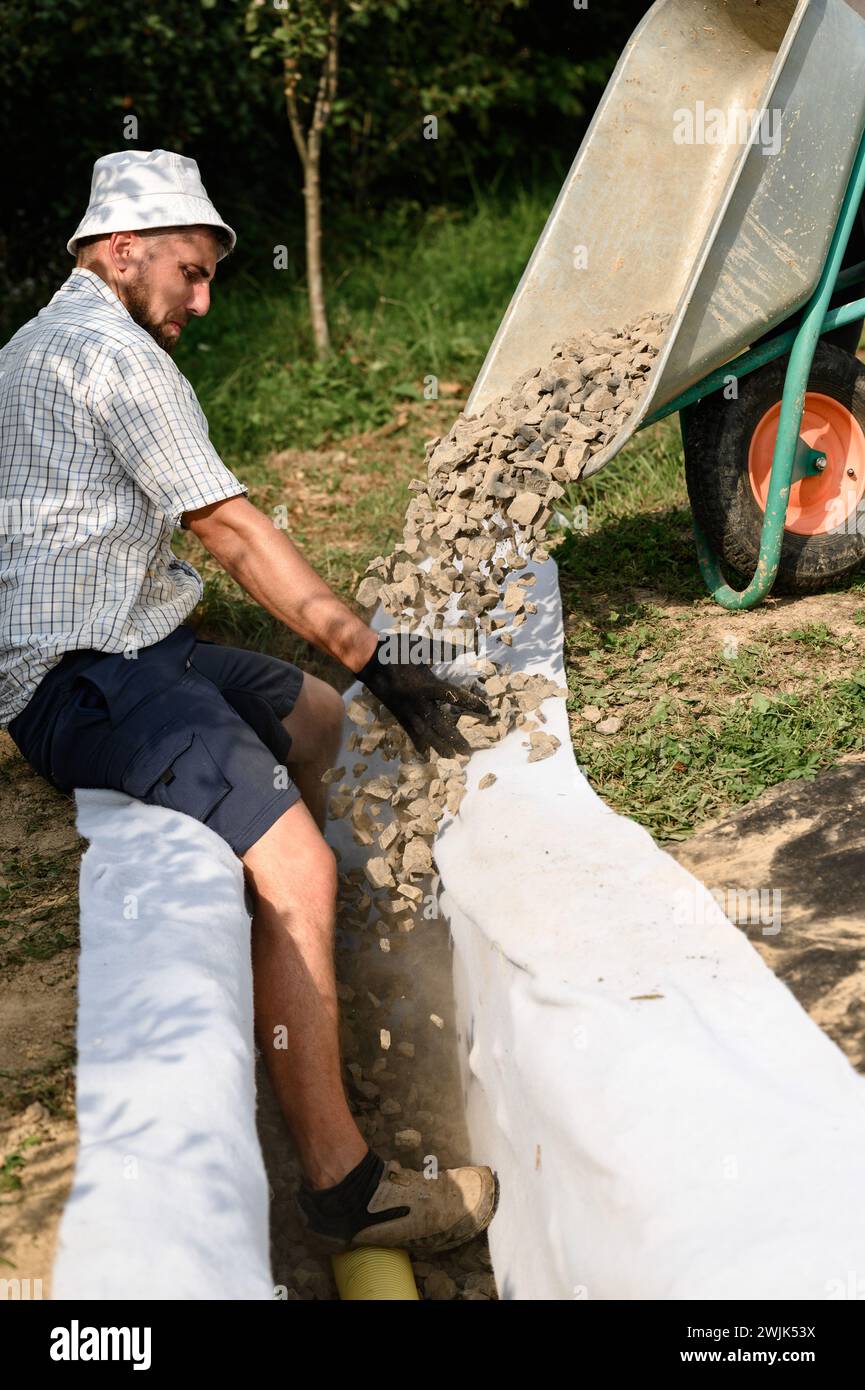 A man pours rubble crushed stone from a wheelbarrow into a trench ...