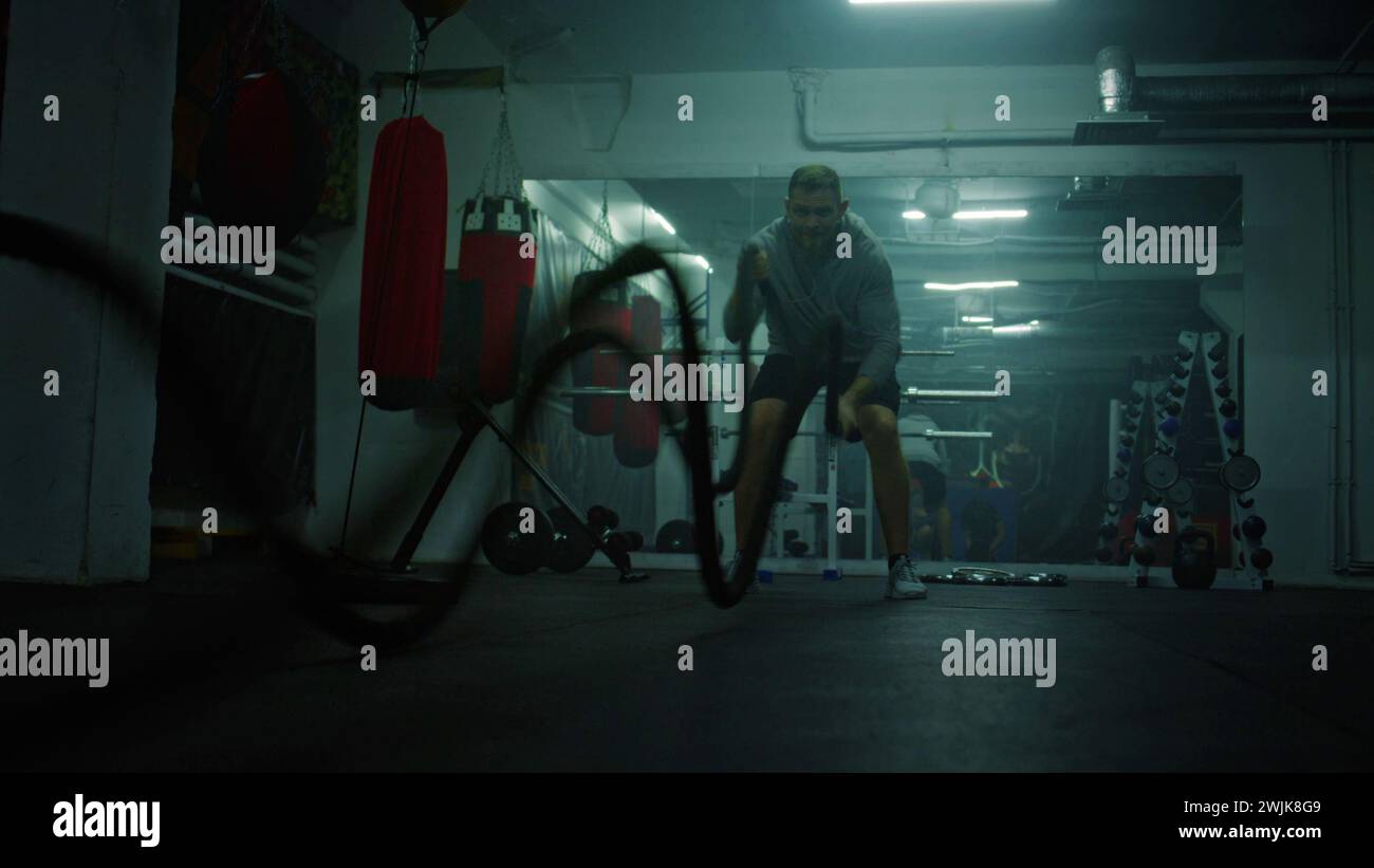 Athletic man exercises with battle ropes in dark boxing gym ...