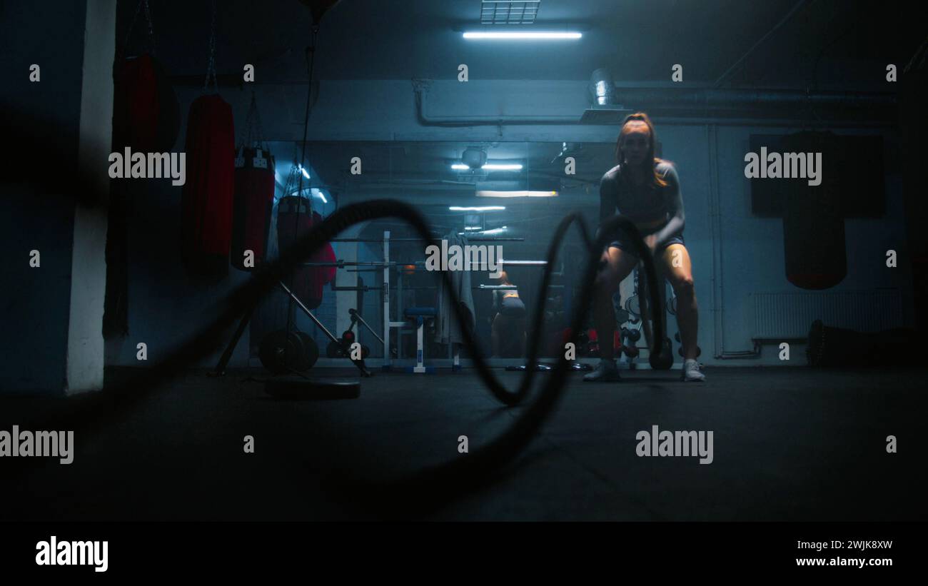 Female athlete exercises with battle ropes in dark boxing gym with LED ...