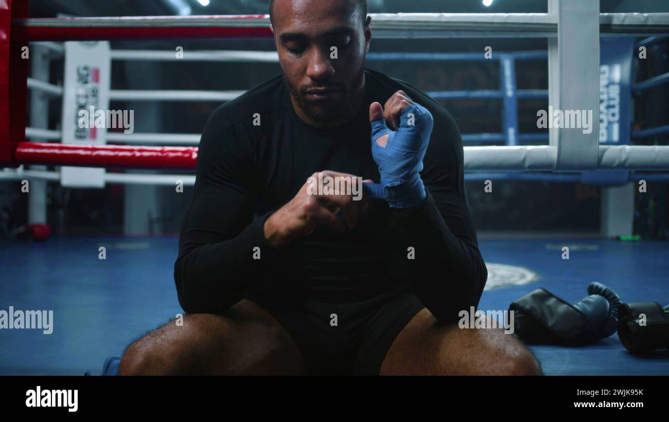African American boxer sits near boxing ring and wraps his hands with ...