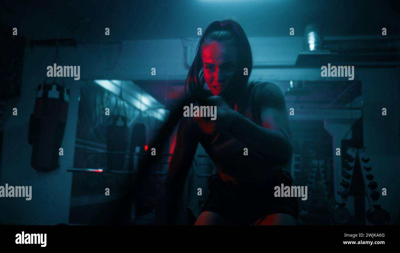 Athletic woman trains with battle ropes in dim boxing gym with LED ...