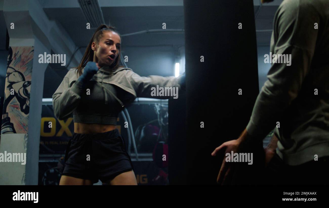 Female fighter in boxing bandages exercises with male trainer in dark ...