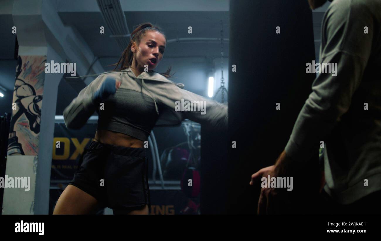 Female fighter in boxing bandages exercises with male trainer in dark ...