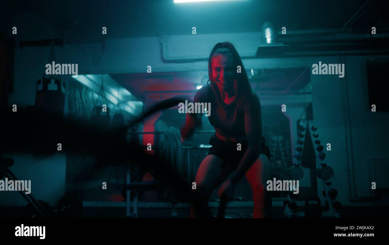 Athletic woman trains with battle ropes in dim boxing gym with LED ...