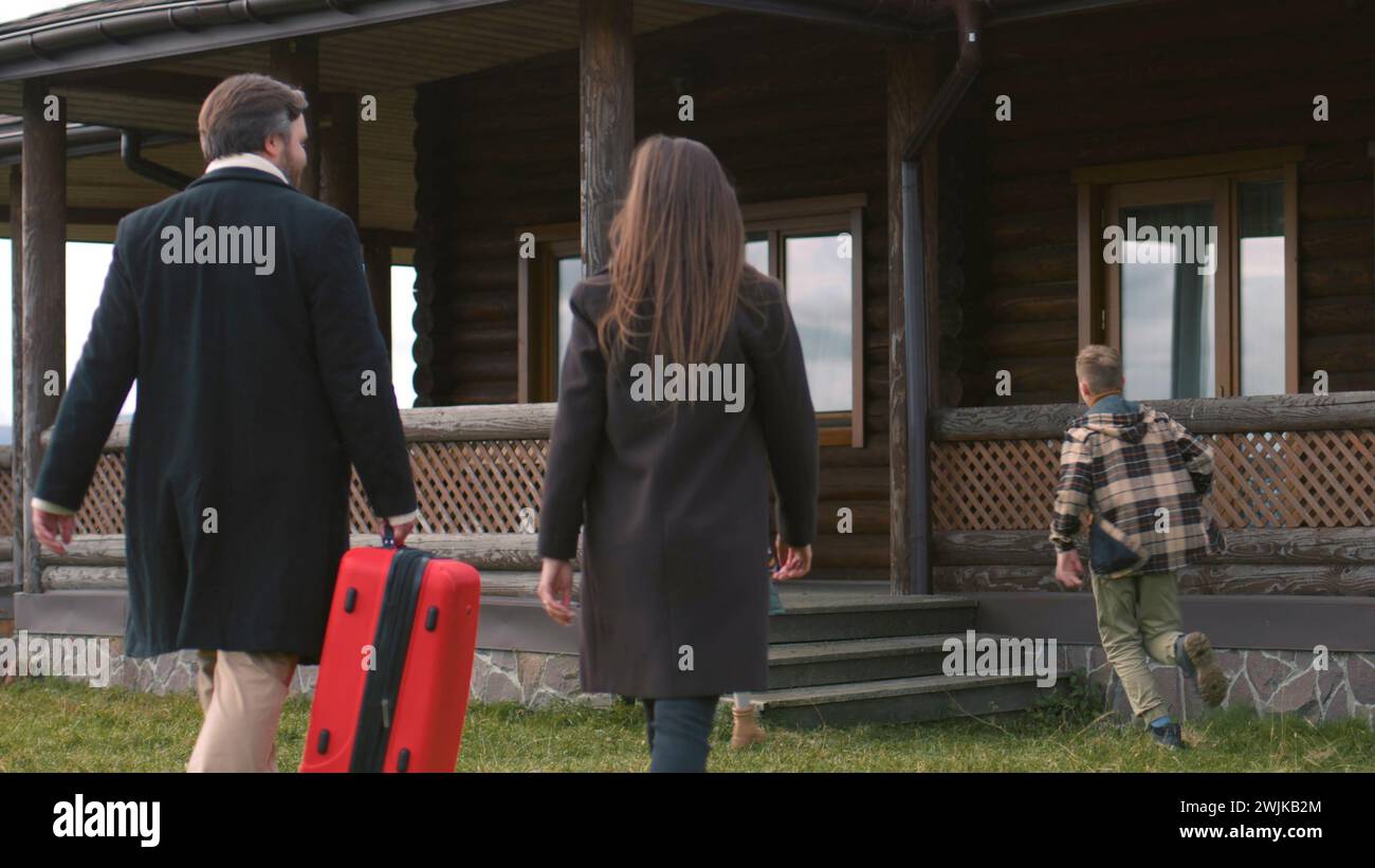 Couple with kids walks to wooden cottage with suitcase. Happy family ...