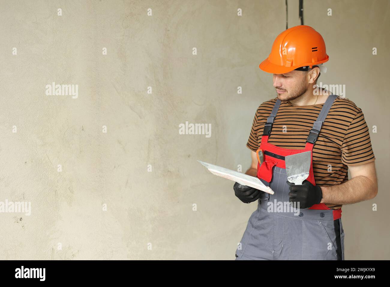 repairman in overalls and protective orange helmet holds two spatulas ...