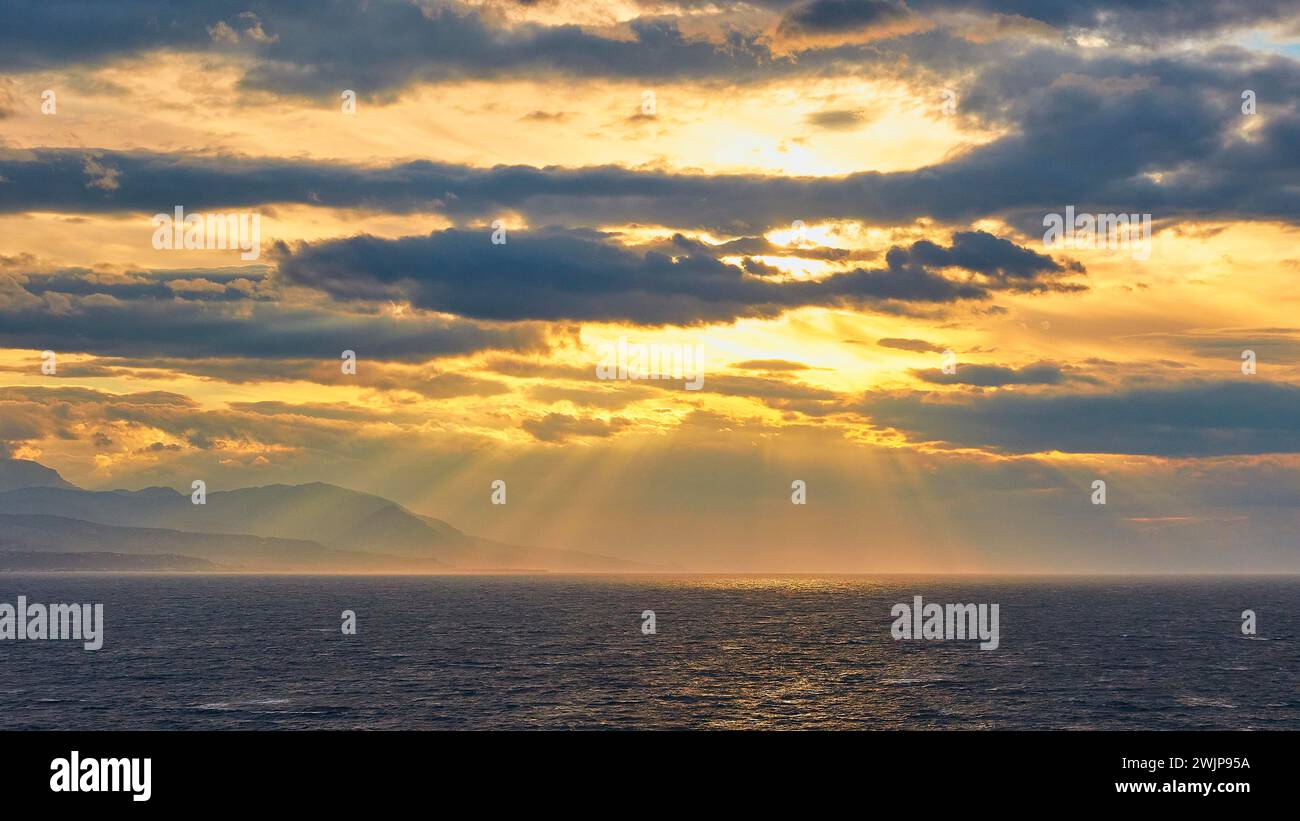 The sun sends its rays through the clouds over the sea near a mountain ...