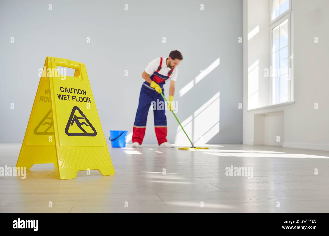 Caution wet floor sign with male young cleaner man mopping empty room ...