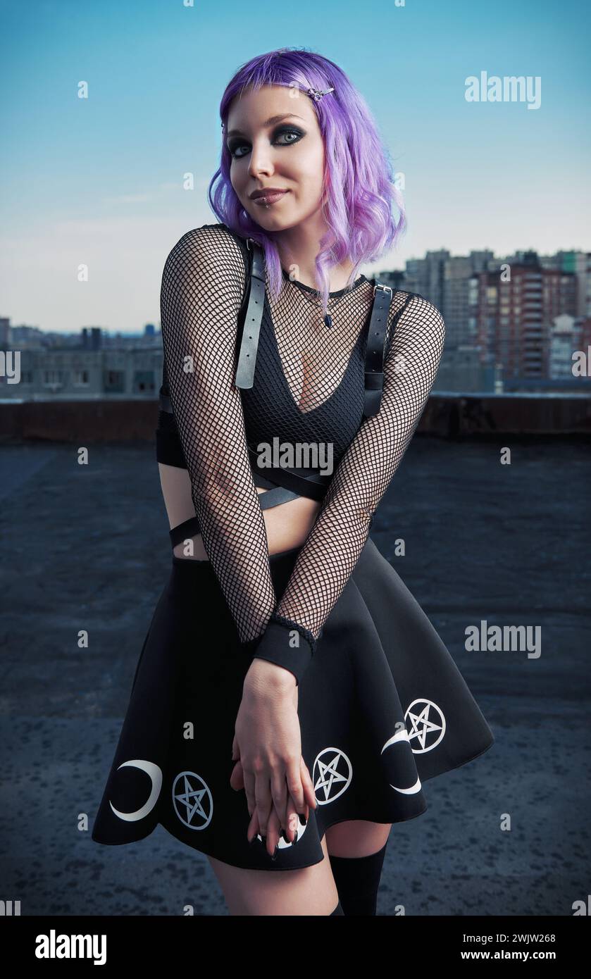 Portrait of cute smiling gothic girl on the roof at evening time. Pastel goth with violet ...