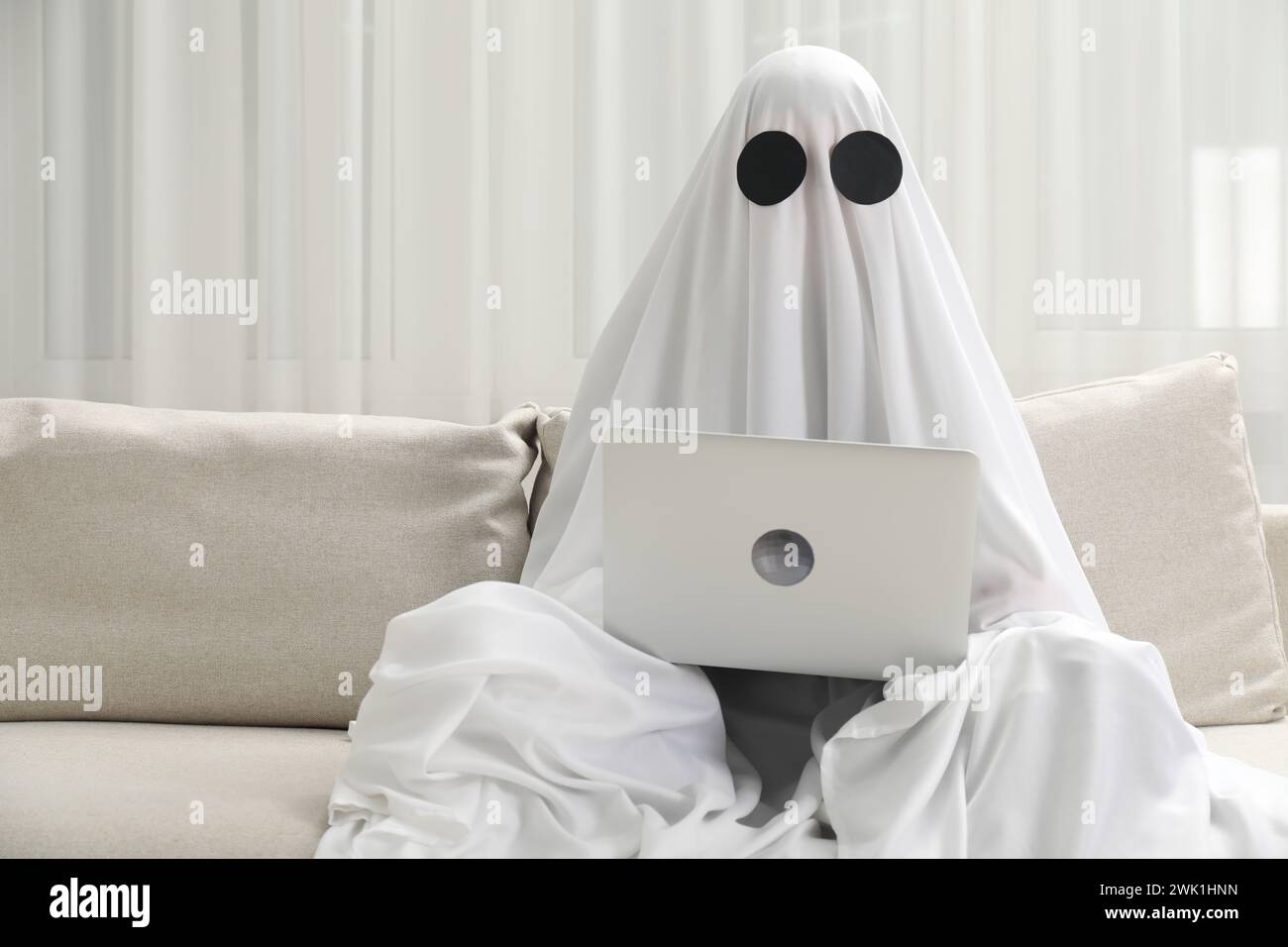 Creepy ghost. Person covered with white sheet using laptop on sofa at ...