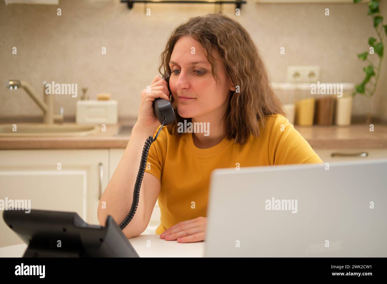 A woman with a laptop is talking on a landline phone at a table in a ...