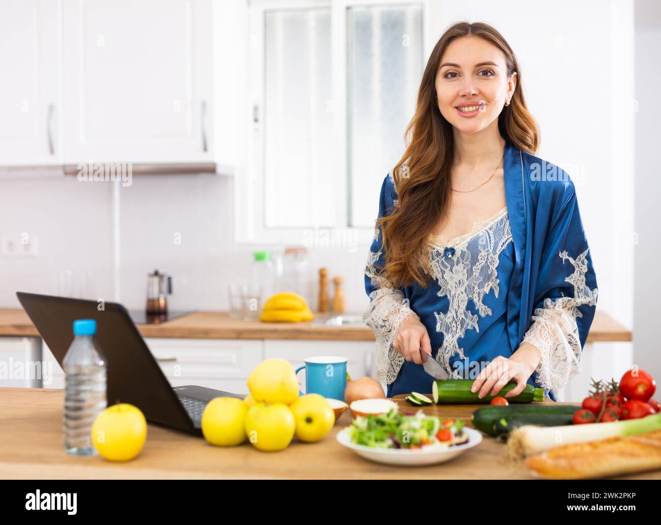 Young woman in blue robe cooking and watching TV series on laptop Stock ...