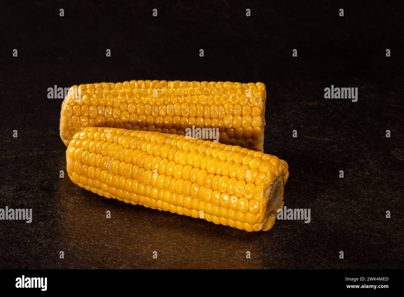 A close-up photo of two fresh ripe corn cobs arranged on a dark black ...