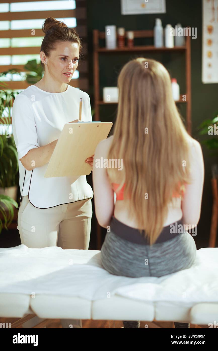 Healthcare time. concerned massage therapist woman in massage cabinet with clipboard and teenage ...