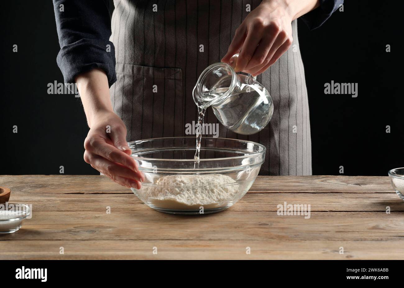 Making bread. Woman pouring water into bowl with flour at wooden table ...