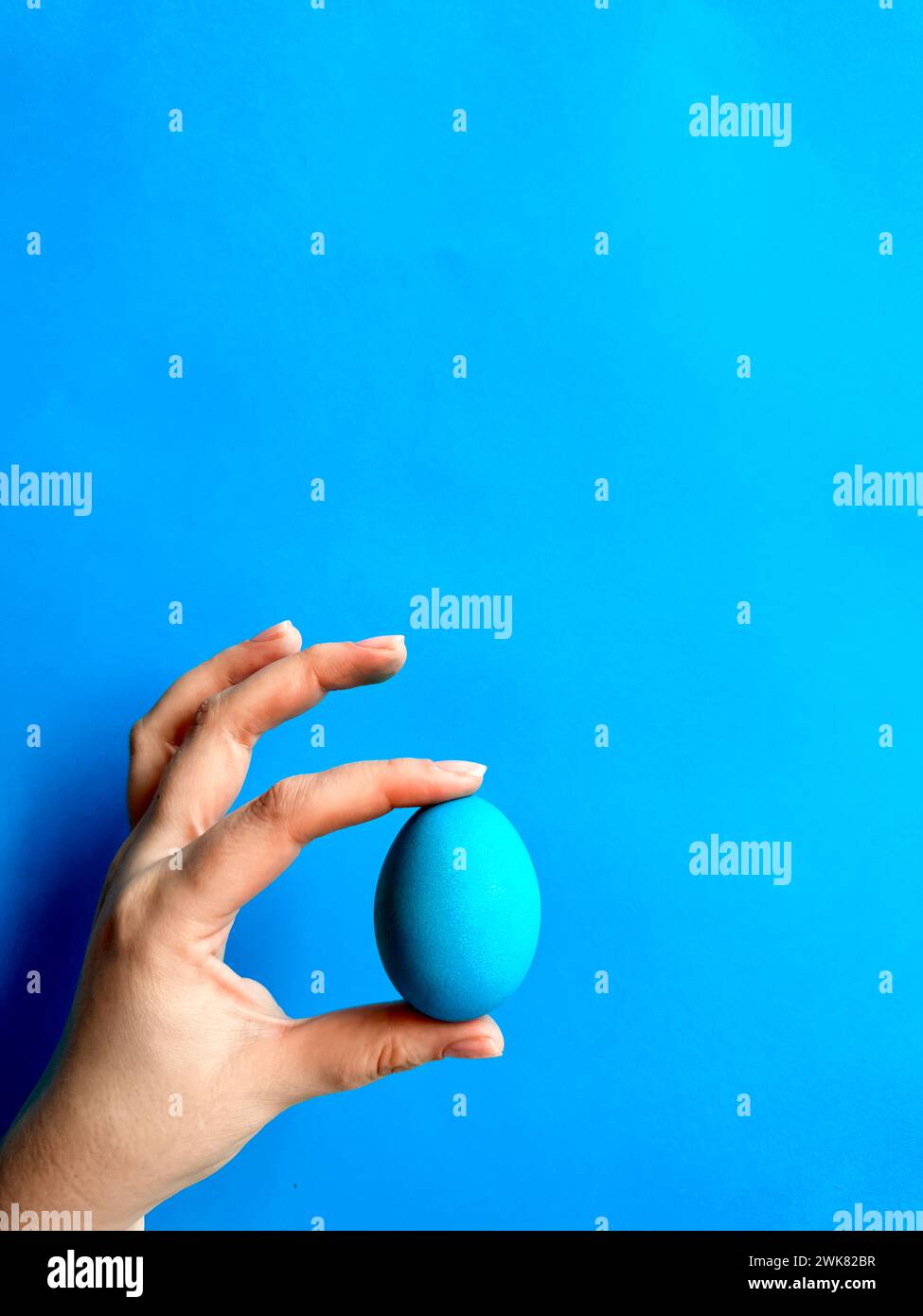Hand balancing blue egg on a fingertip against solid blue background ...