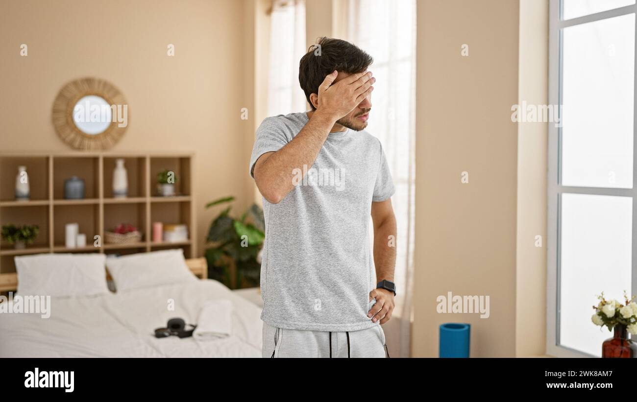 A young hispanic man with a beard stands distressed in a bedroom ...