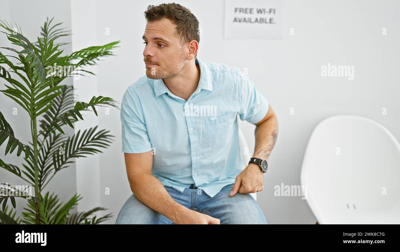 Hispanic man sitting casually in an indoor white lobby with a potted ...