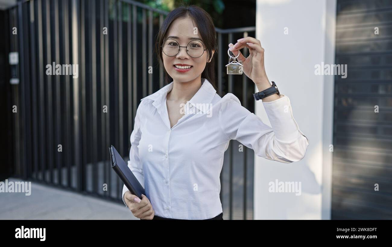 A young asian woman realtor smilingly displays house keys on an urban ...