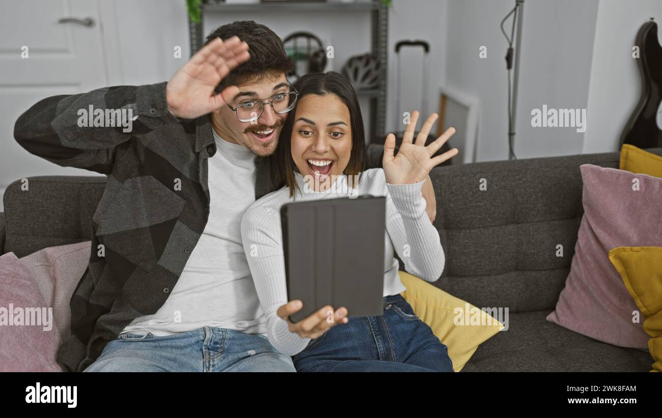 A cheerful couple waves hello during a video call in their cozy living ...