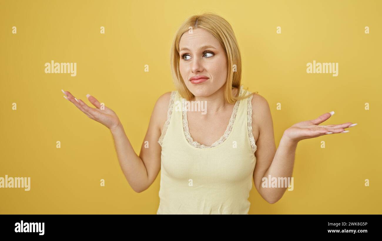 A perplexed young blonde woman shrugging against a yellow background ...