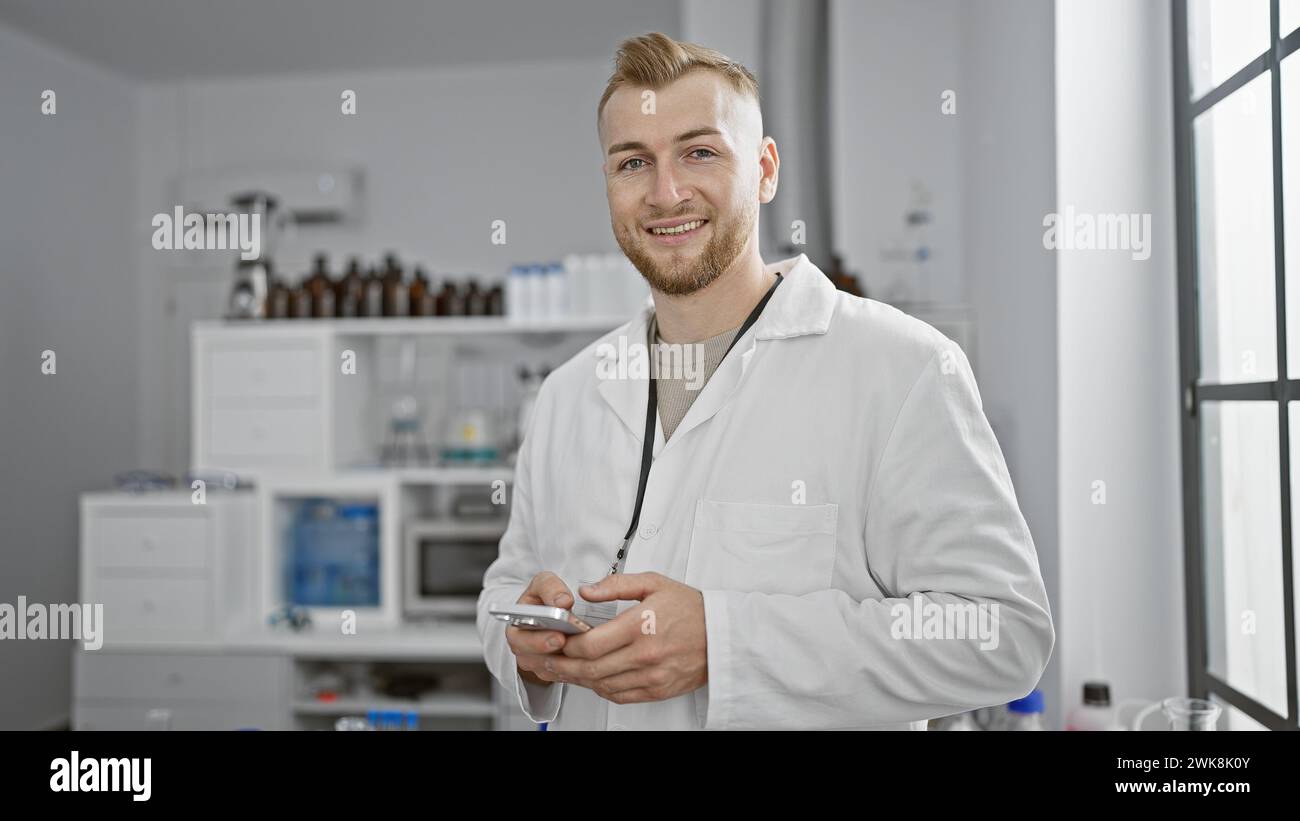 A young, bearded man in a white lab coat holding a smartphone smiles ...