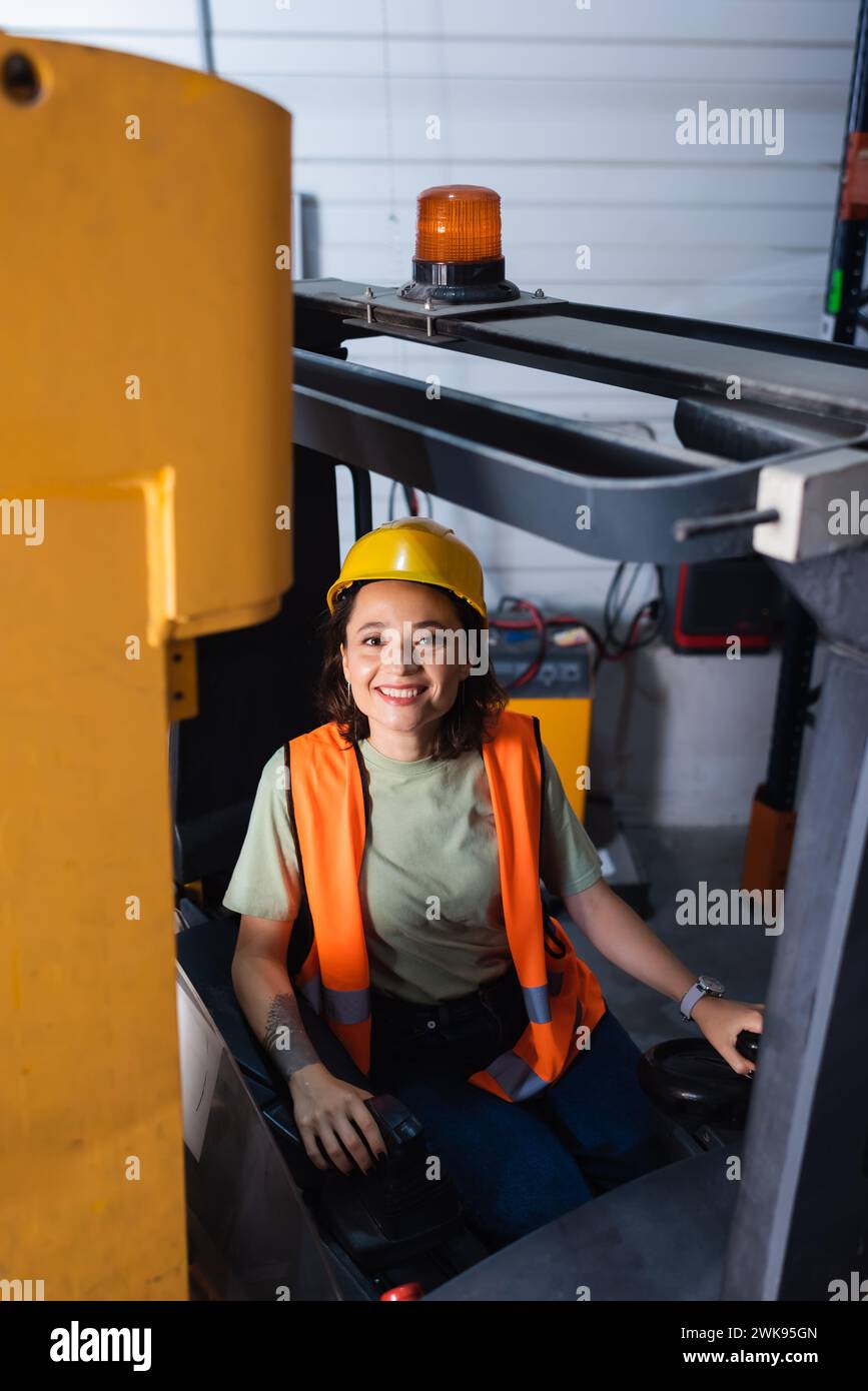 cheerful female forklift operator in hard hat and safety vest looking ...
