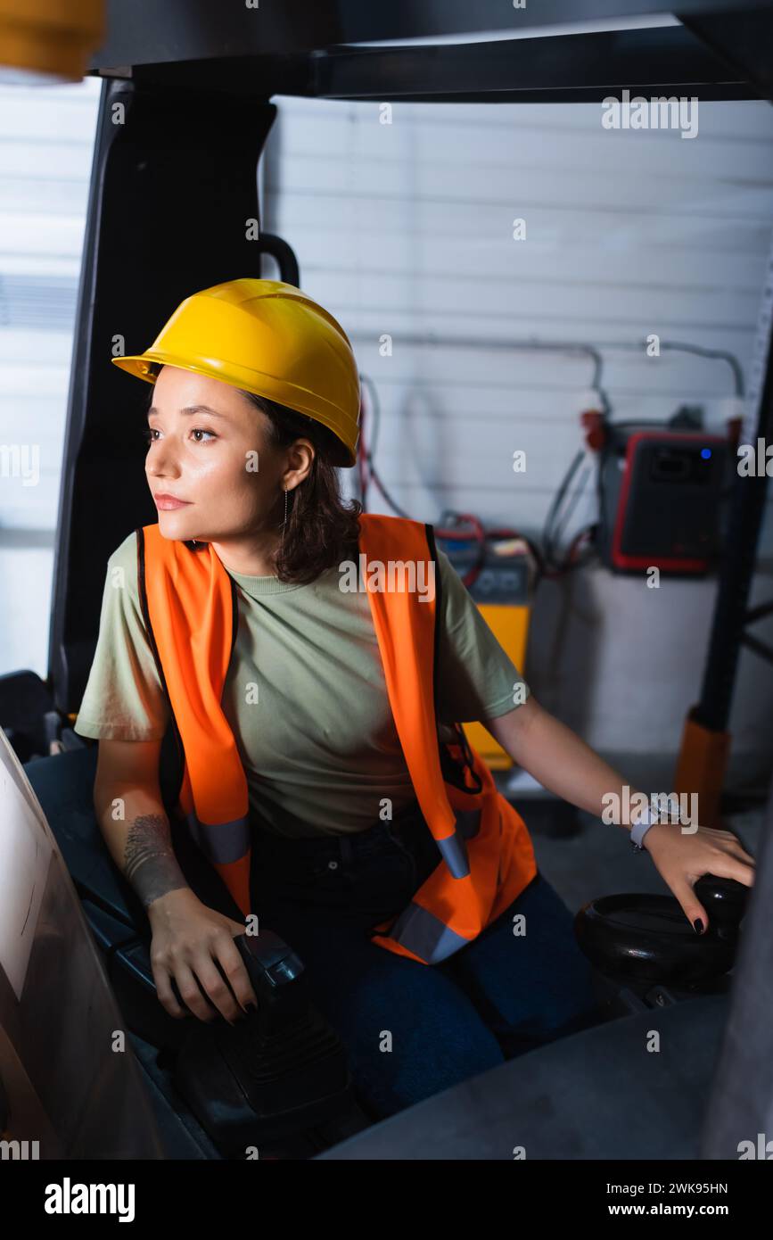 pensive female forklift operator in hard hat and safety vest looking ...
