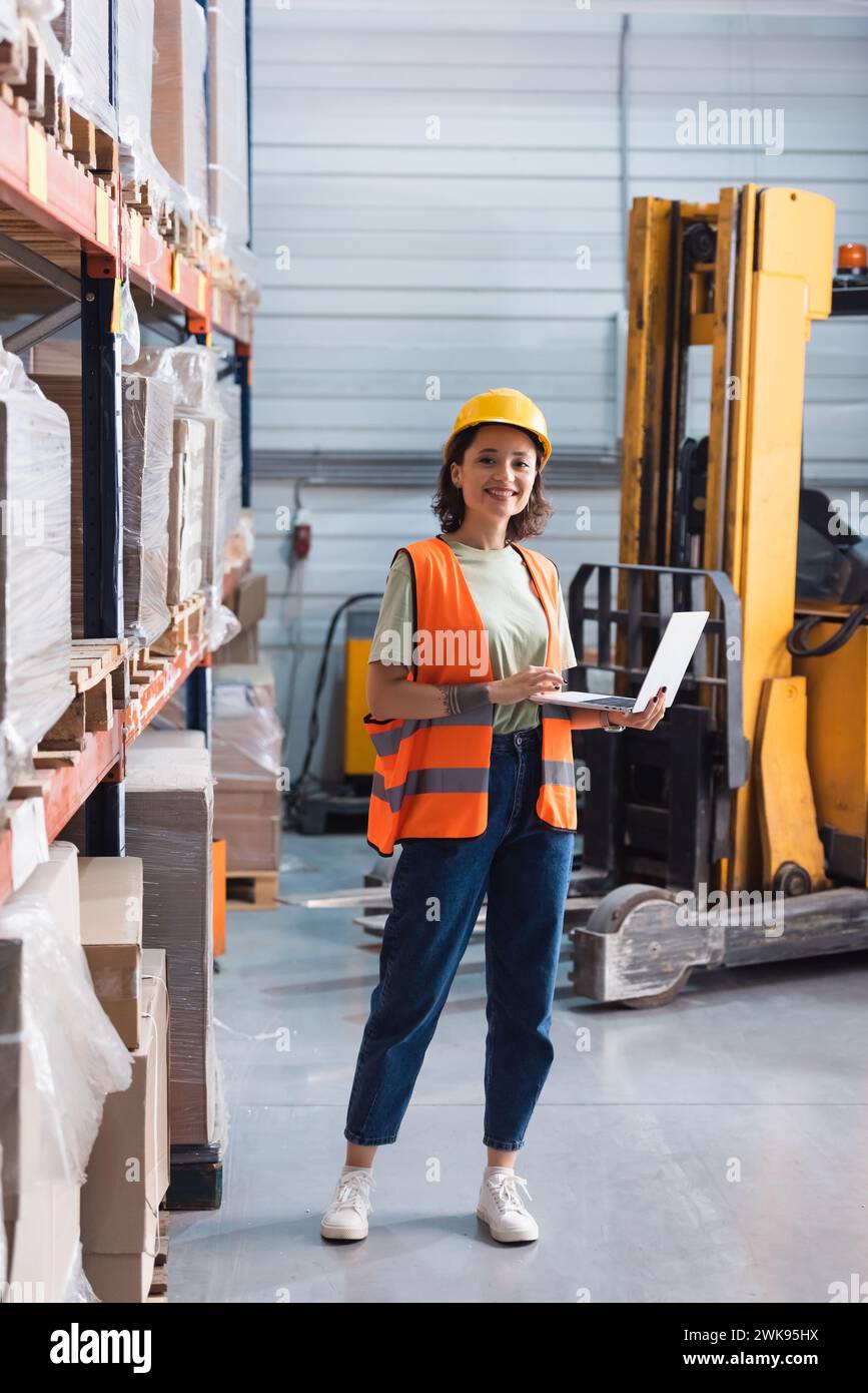 happy warehouse female worker in hard hat and safety vest using laptop while checking inventory ...