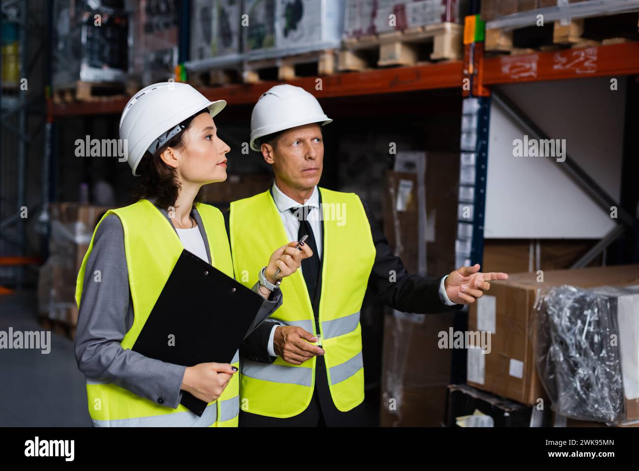 Warehouse supervisor instructing female employee in hard hat and safety ...