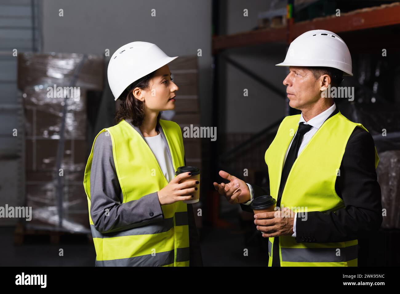 logistics team, woman in hard hat and coffee chatting with middle aged ...