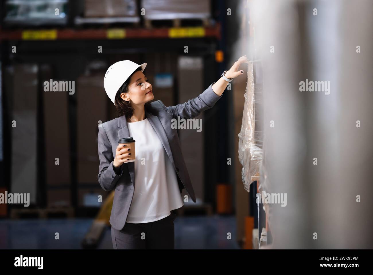 businesswoman in suit and heard hat holding coffee to go in warehouse ...