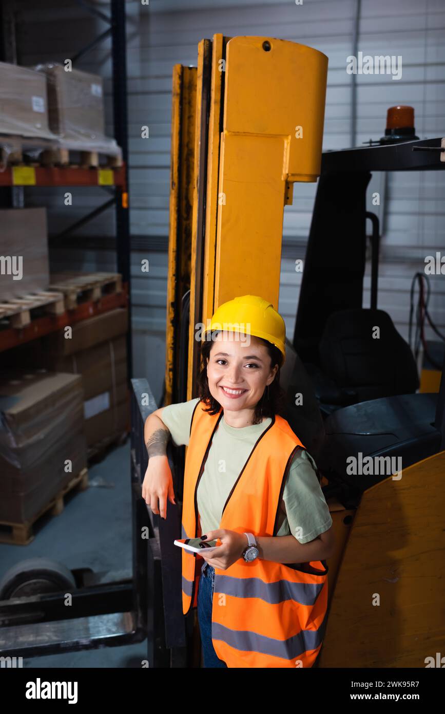 happy female warehouse worker in hard hat and safety vest holding smartphone near forklift ...