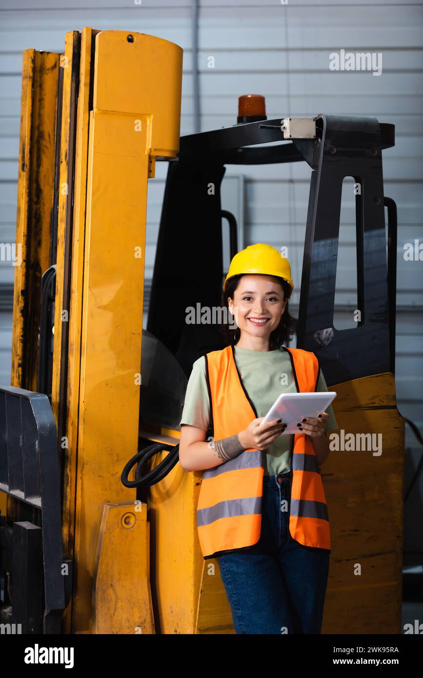 happy female warehouse worker in hard hat and safety vest holding tablet near forklift, cargo ...