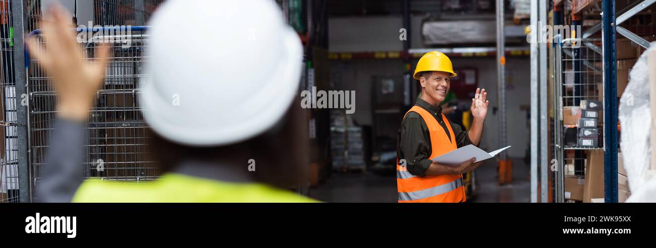 happy supervisor in hard hat waving at colleague while greeting each ...