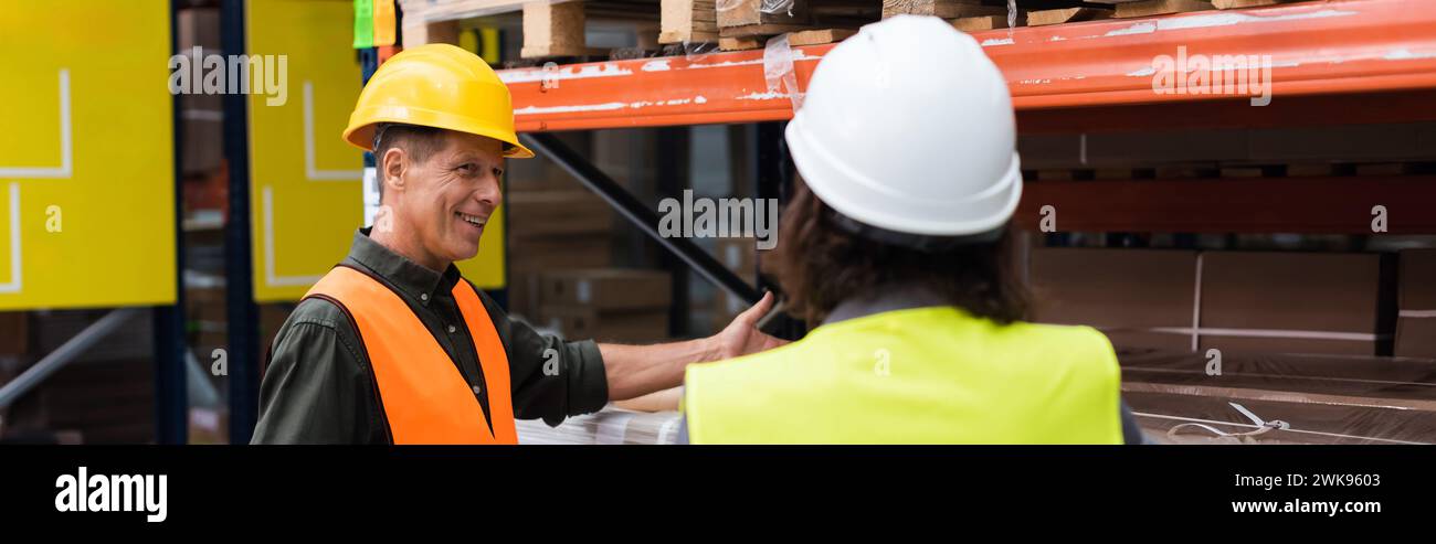 happy middle aged supervisor in hard hat gesturing and explaining work to warehouse employee ...
