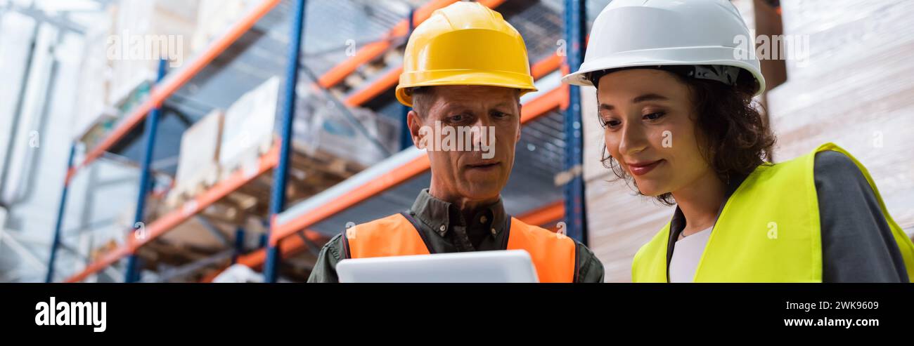happy supervisor in hard hat holding tablet while explaining work to employee in warehouse ...