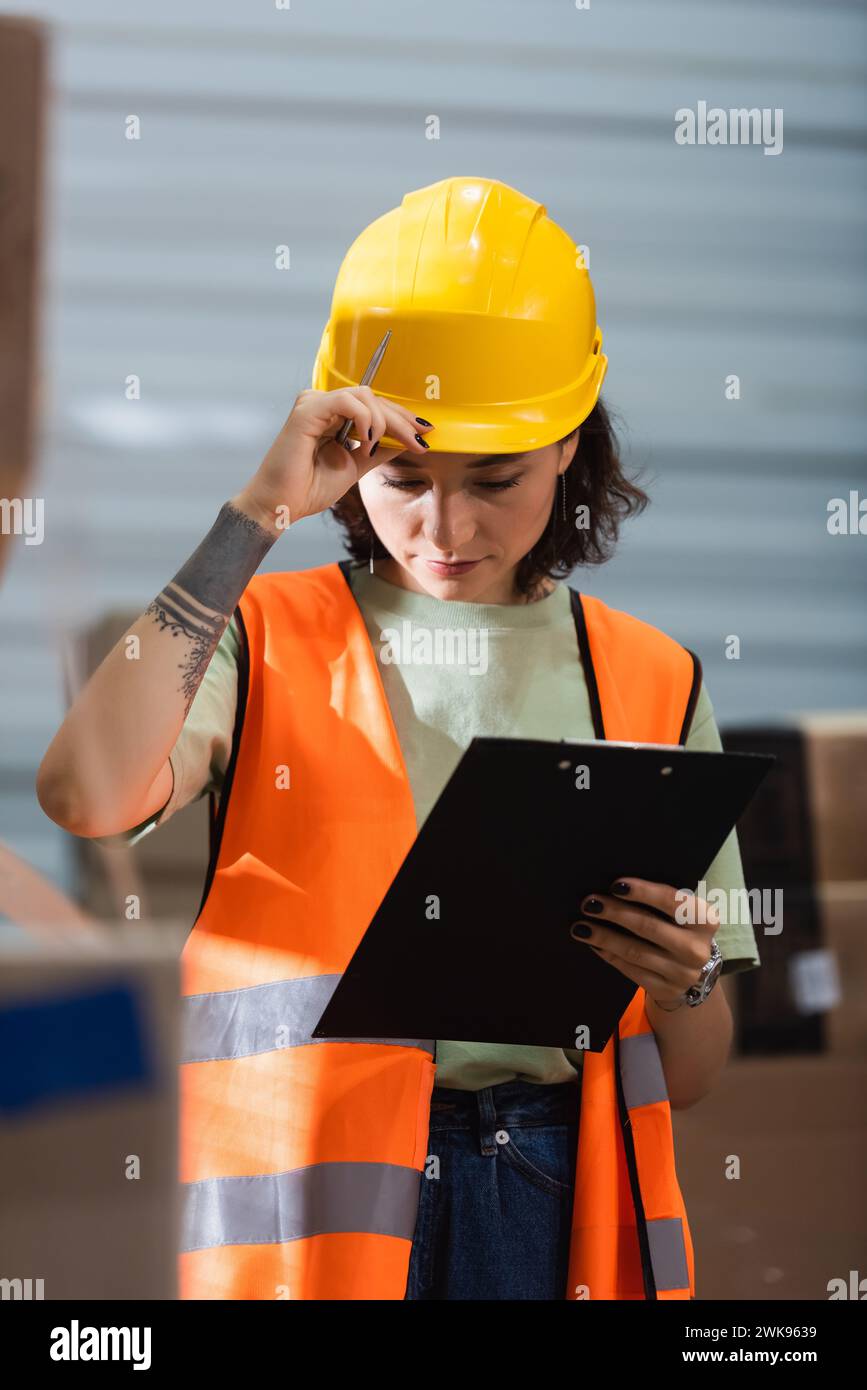 female warehouse worker in safety vest adjusting hard hat and holding ...