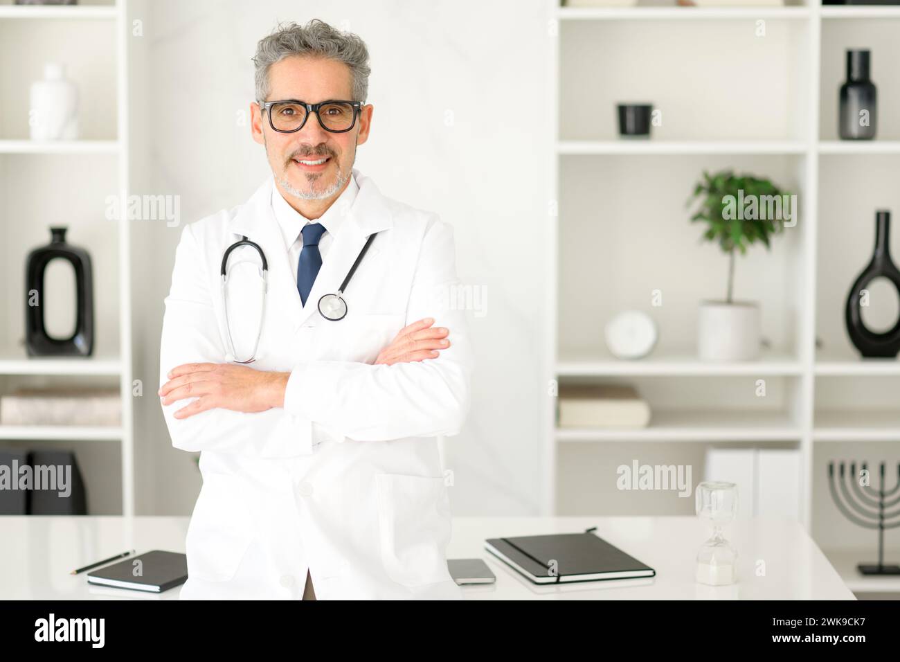 Senior doctor stands with arms crossed in office, indicating a blend of ...