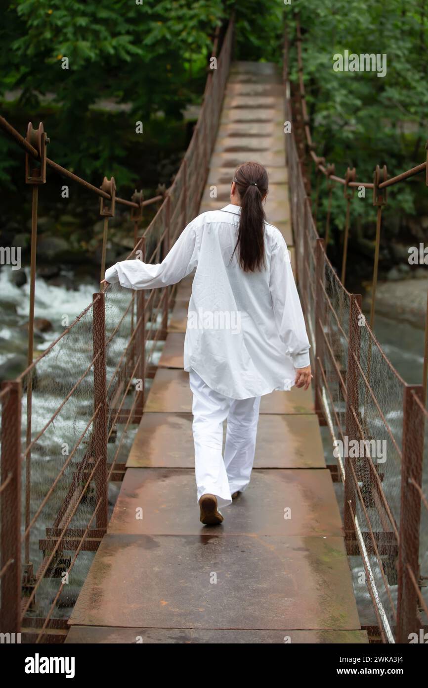 A woman in white clothes walks backwards along a suspension bridge over ...