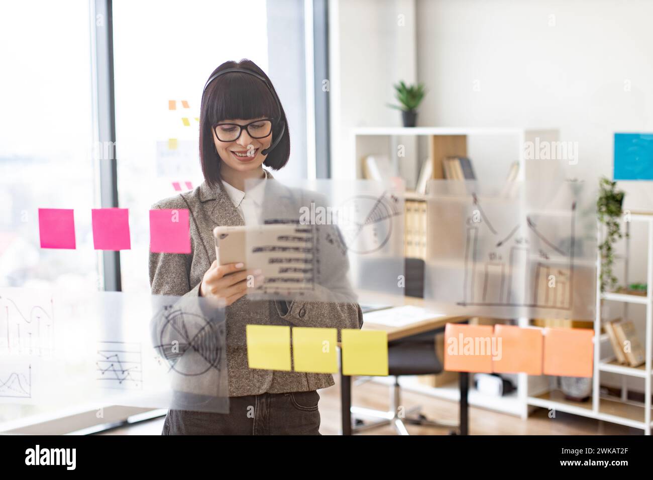 Focused young female standing near glass wall and taking notes during ...